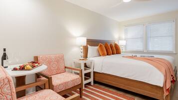 Quarto de hotel com uma cama, uma mesinha com uma travessa de frutas, duas cadeiras, uma mesinha de cabeceira com abajur e uma janela com persianas.