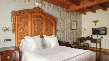 Quarto de hotel com uma grande cabeceira de madeira, cama com roupa de cama branca, escrivaninha com cadeira e televisão na parede.