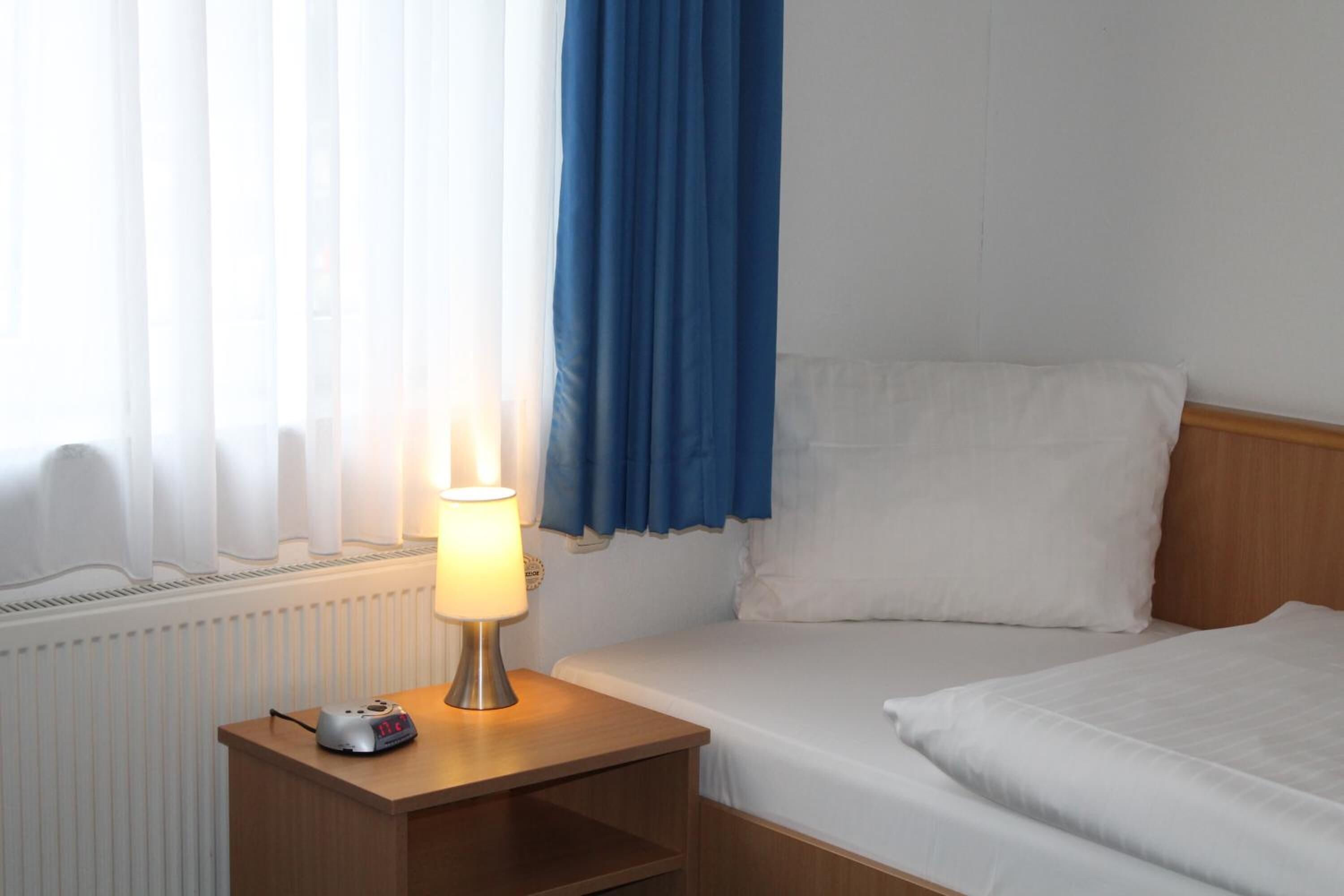 Quarto de hotel com cama, mesa de cabeceira com luminária, rádio e janela com cortinas.