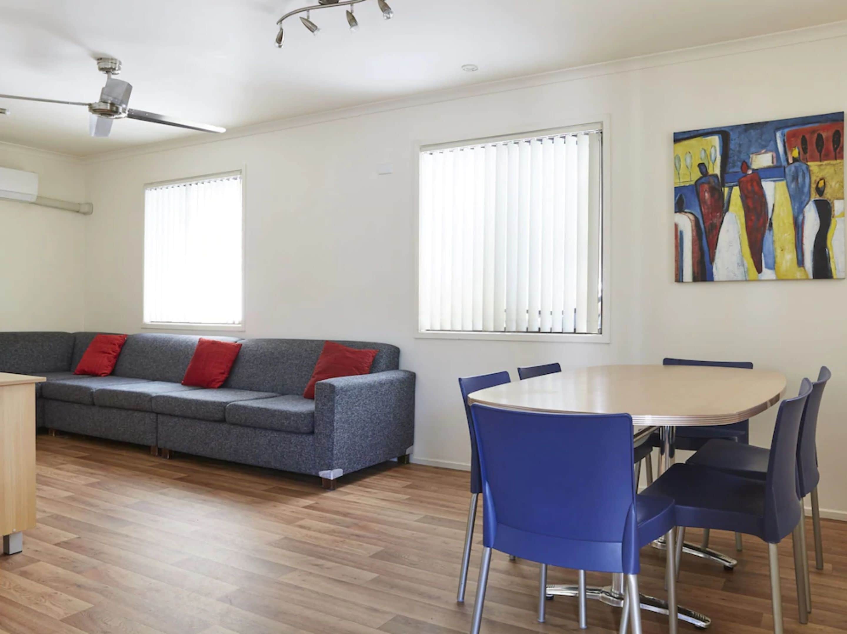Uma sala de estar moderna com um sofá cinzento, almofadas vermelhas, uma mesa de jantar de madeira com cadeiras azuis e um ventilador de teto.