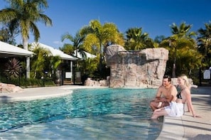 Uma área de piscina com água cristalina, uma formação rochosa e palmeiras.