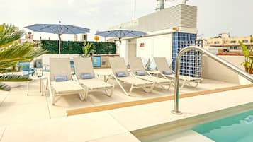 Uma área de piscina no terraço, com espreguiçadeiras, guarda-sóis e um pequeno edifício com fachada azul.