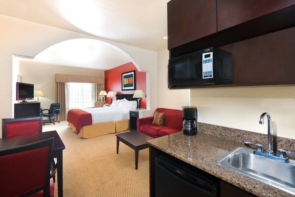Quarto de hotel com cama, sofá vermelho, escrivaninha, cafeteira e cozinha equipada com pia e micro-ondas.