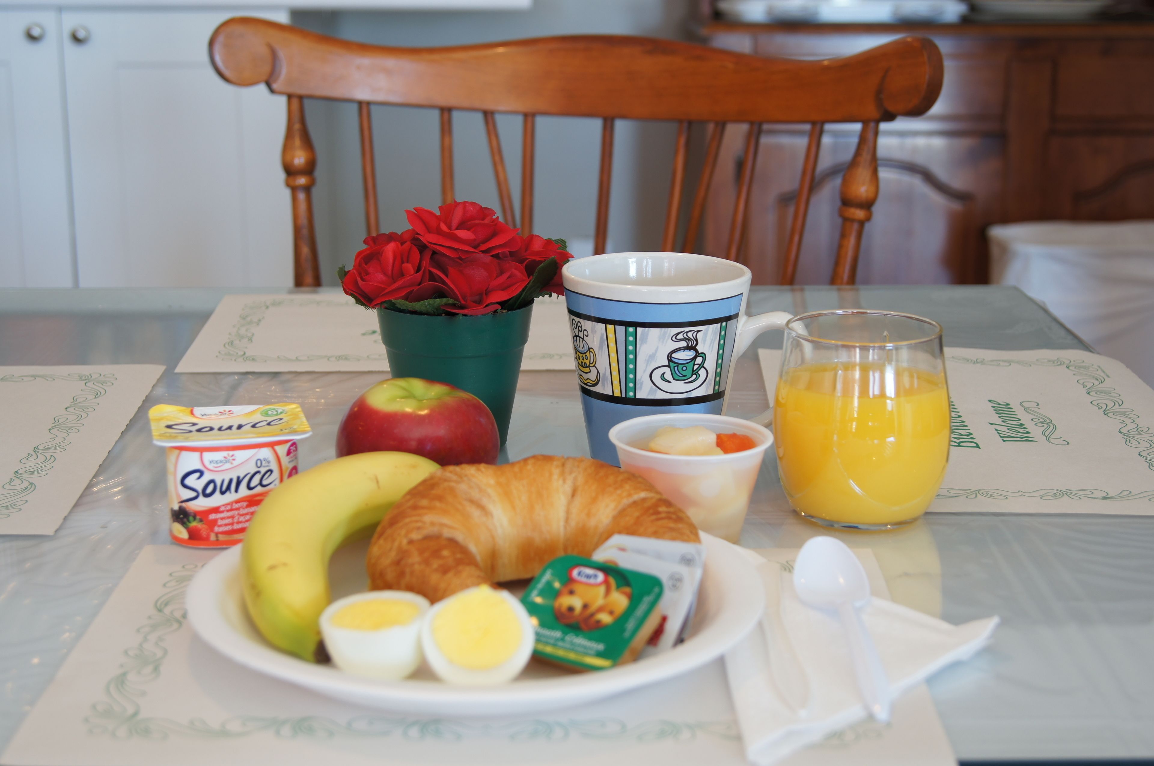 Mesa de café da manhã com croissant, maçã, banana, ovos, iogurte, copo de suco de laranja e caneca com personagem de desenho animado.