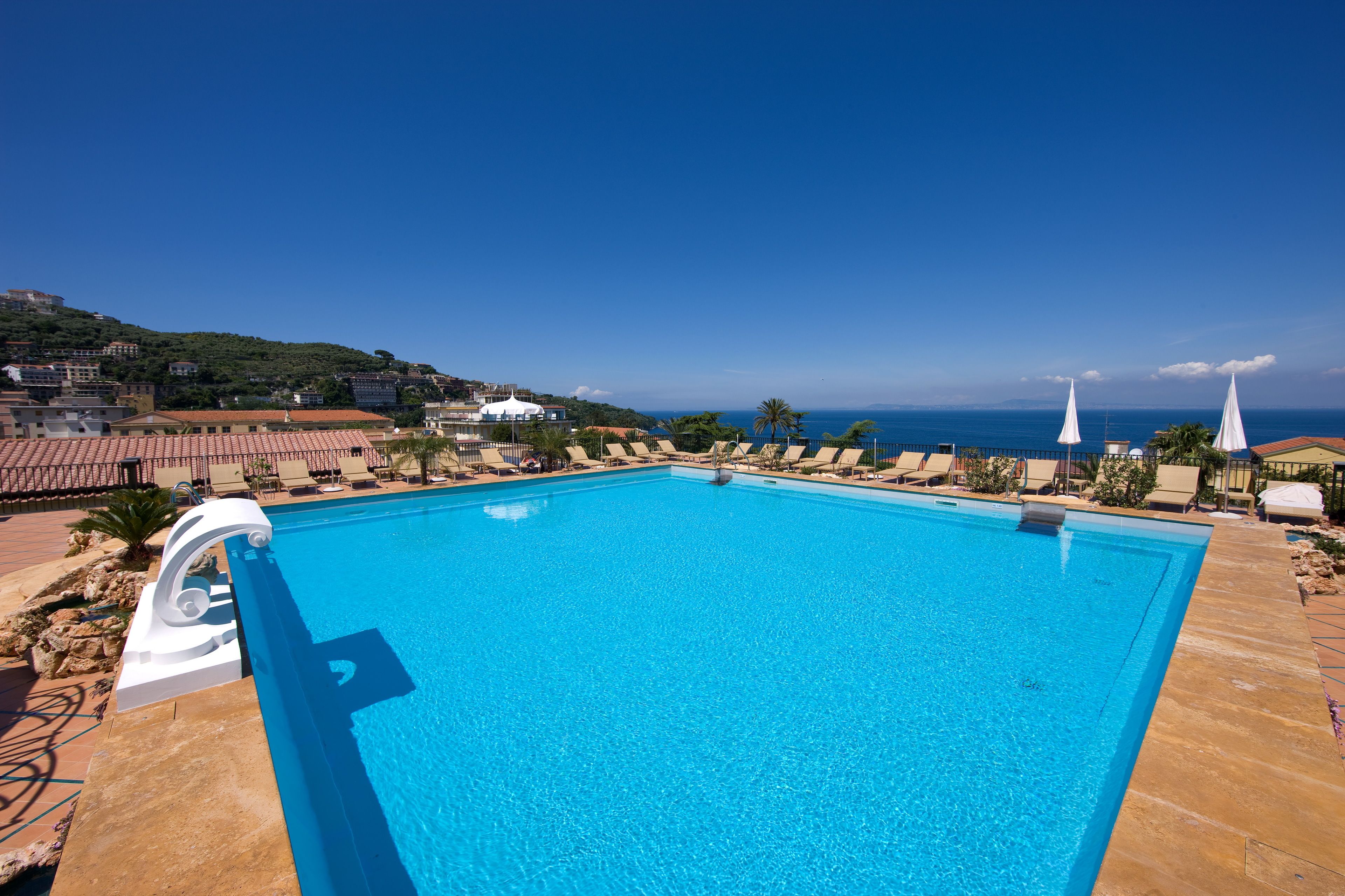 Uma piscina retangular com águas azuis cristalinas, rodeada por espreguiçadeiras e guarda-sóis, com vista para o mar e colinas ao fundo.
