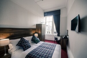 Quarto de hotel com cabeceira de madeira, cama com roupa de cama xadrez, luminária de cabeceira, escrivaninha com cadeira, televisão montada na parede e janela com cortinas.