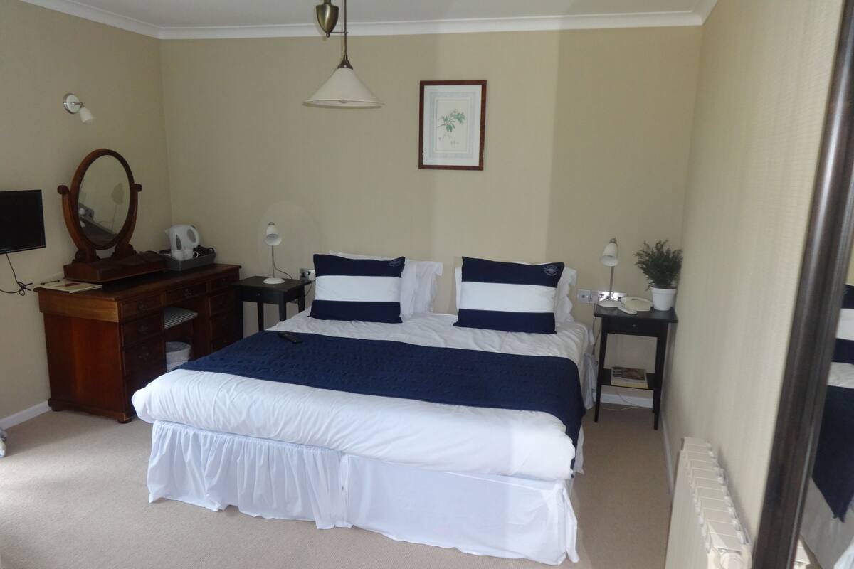 Quarto com cama grande, mesas de cabeceira, penteadeira, espelho, quadro na parede e radiador.