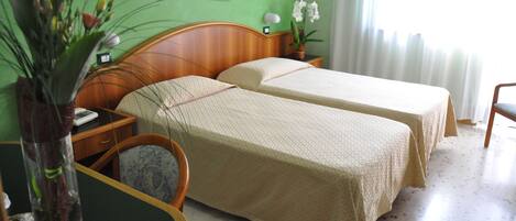 Quarto de hotel com duas camas, cabeceira de madeira, mesa de cabeceira com abajur, uma cadeira, escrivaninha e um vaso com flores.