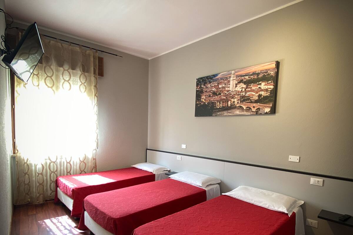 Quarto de hotel com três camas de solteiro, um quadro de uma paisagem urbana, televisão de tela plana e uma janela com cortinas.