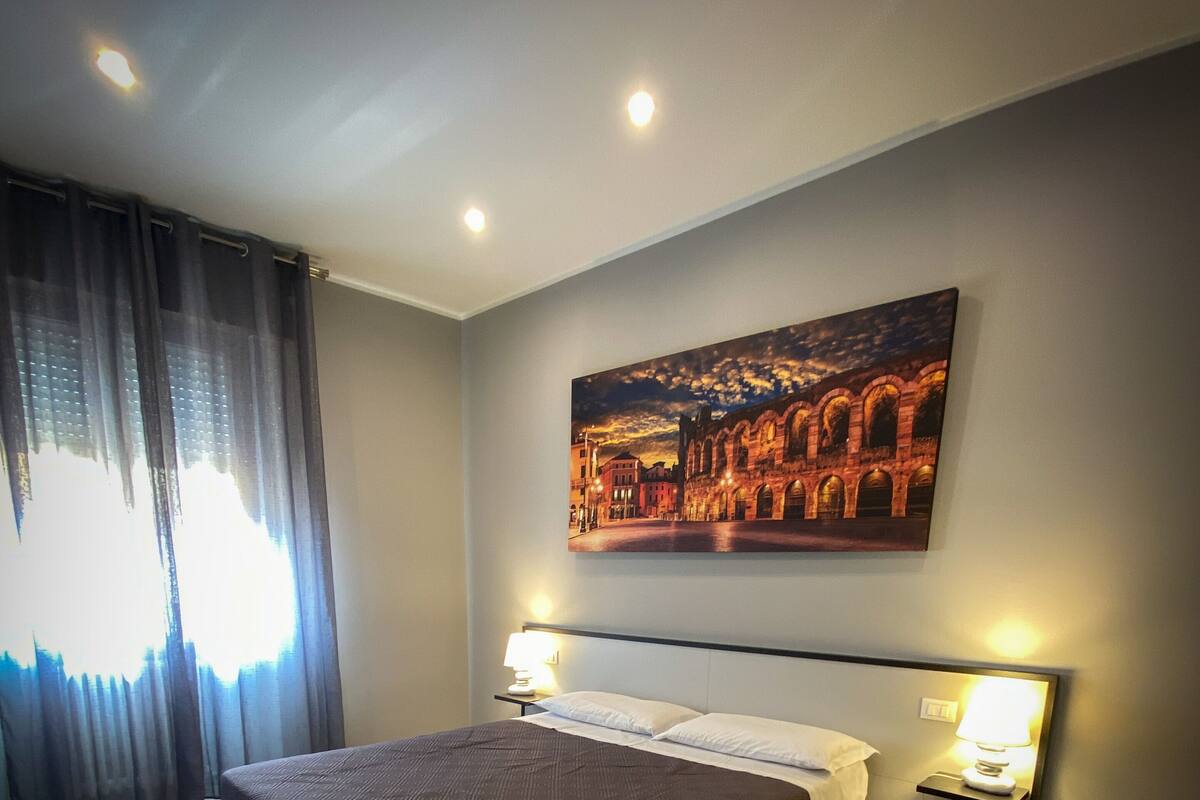 Quarto moderno com uma cama grande, duas mesas de cabeceira com luminárias, uma luz no teto e uma pintura de paisagem urbana na parede.