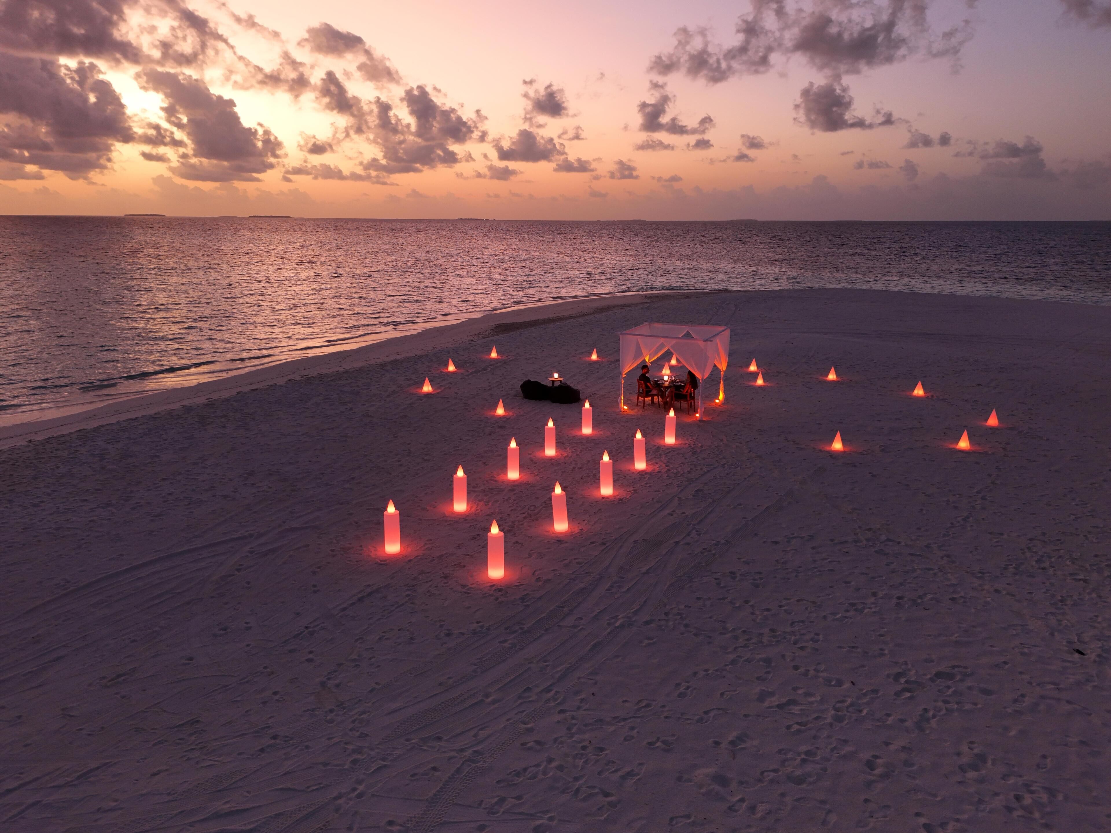 Praia ao pôr do sol, com uma barraca e pessoas reunidas em volta de uma mesa com velas.