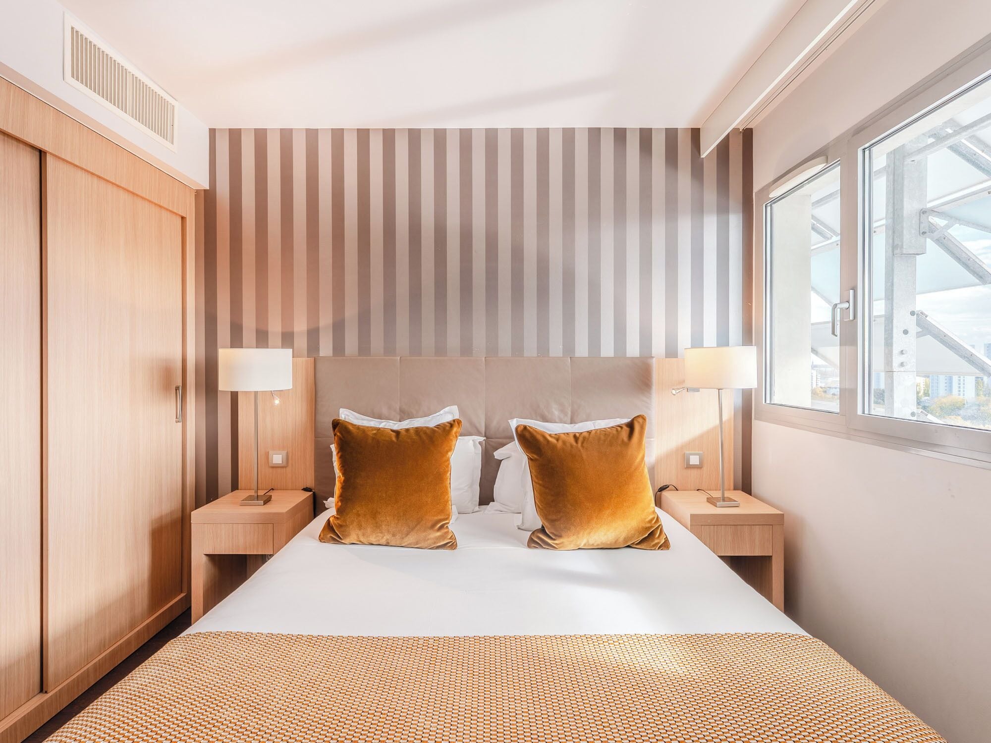 Quarto de hotel com cama grande, duas mesas de cabeceira, parede listrada e janela com vista.