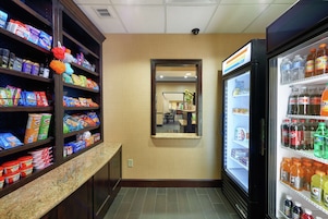 Quarto de hotel com minibar, frigorífico cheio de bebidas e prateleiras com snacks e produtos de higiene pessoal.
