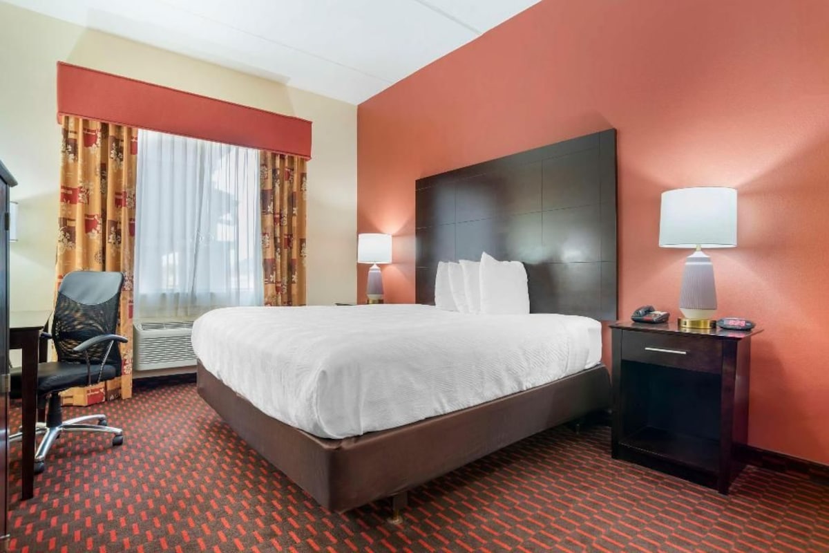 Quarto de hotel com cama, escrivaninha, cadeira e mesa de cabeceira com luminárias.