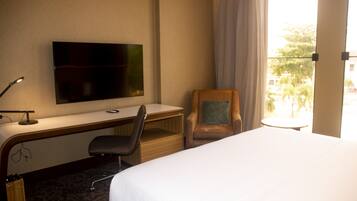 Quarto de hotel com uma cama, uma escrivaninha com cadeira, televisão e janela com cortinas.