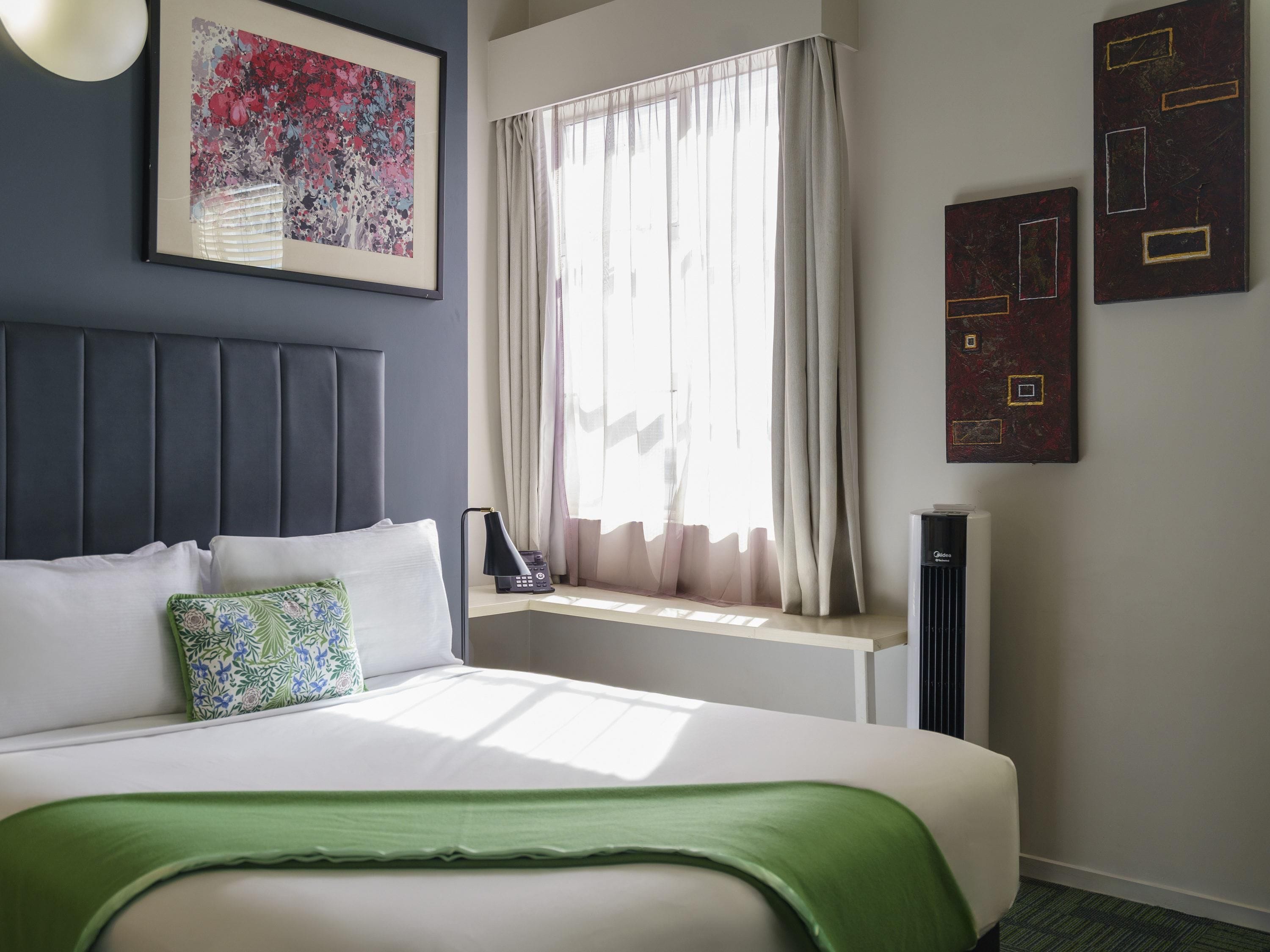 Quarto de hotel com cama, janela com cortinas, radiador e uma obra de arte emoldurada na parede.