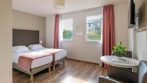 Quarto de hotel com duas camas, uma mesinha com uma planta e uma janela com cortinas.