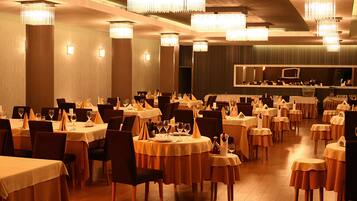 Uma sala de banquetes com várias mesas redondas, cada uma com toalhas brancas e talheres, e grandes lustres pendurados no teto.