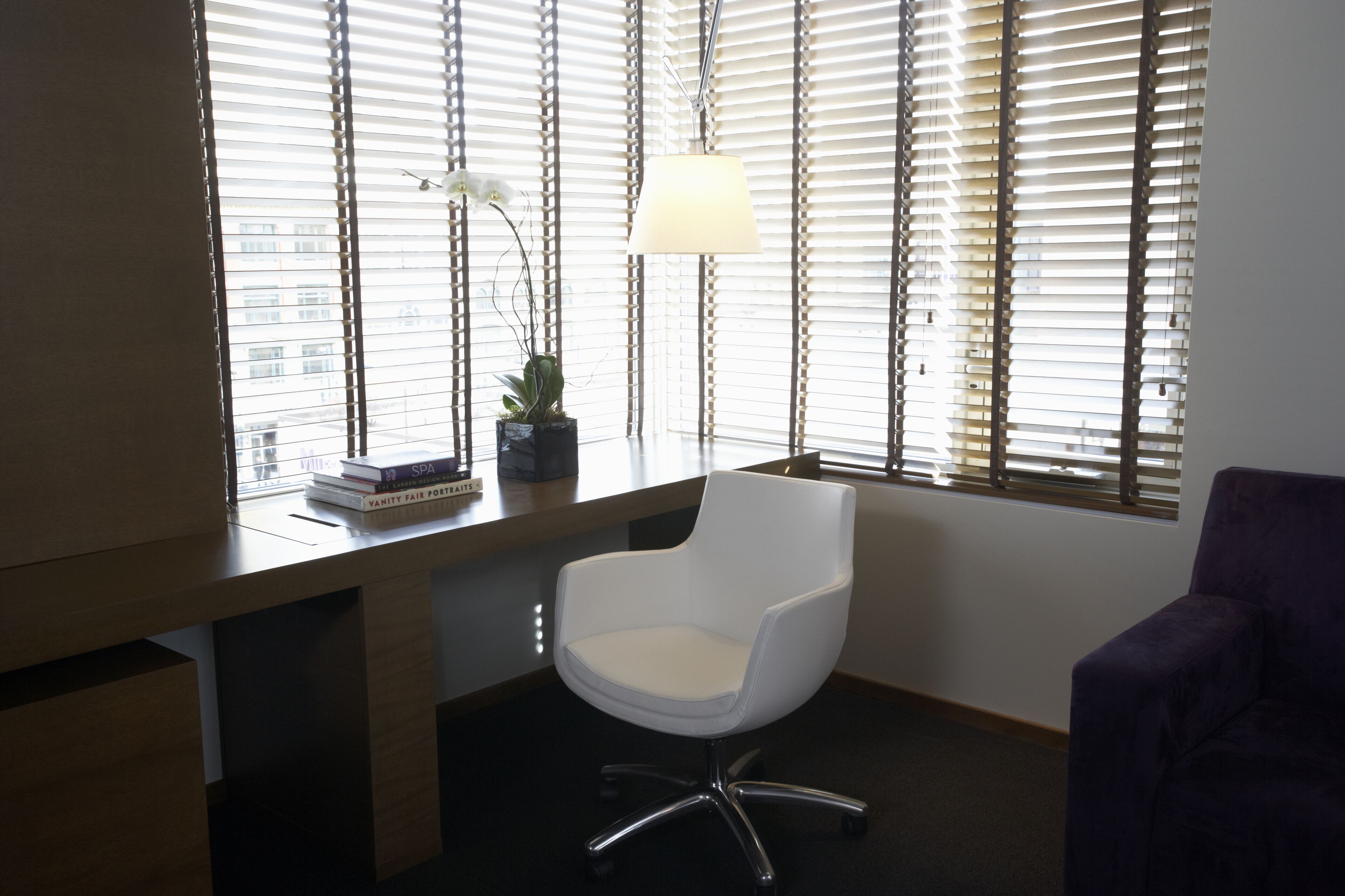 Uma cadeira de escritório moderna com assento branco e pés cromados, posicionada em frente a uma mesa com uma luminária e um vaso de planta.