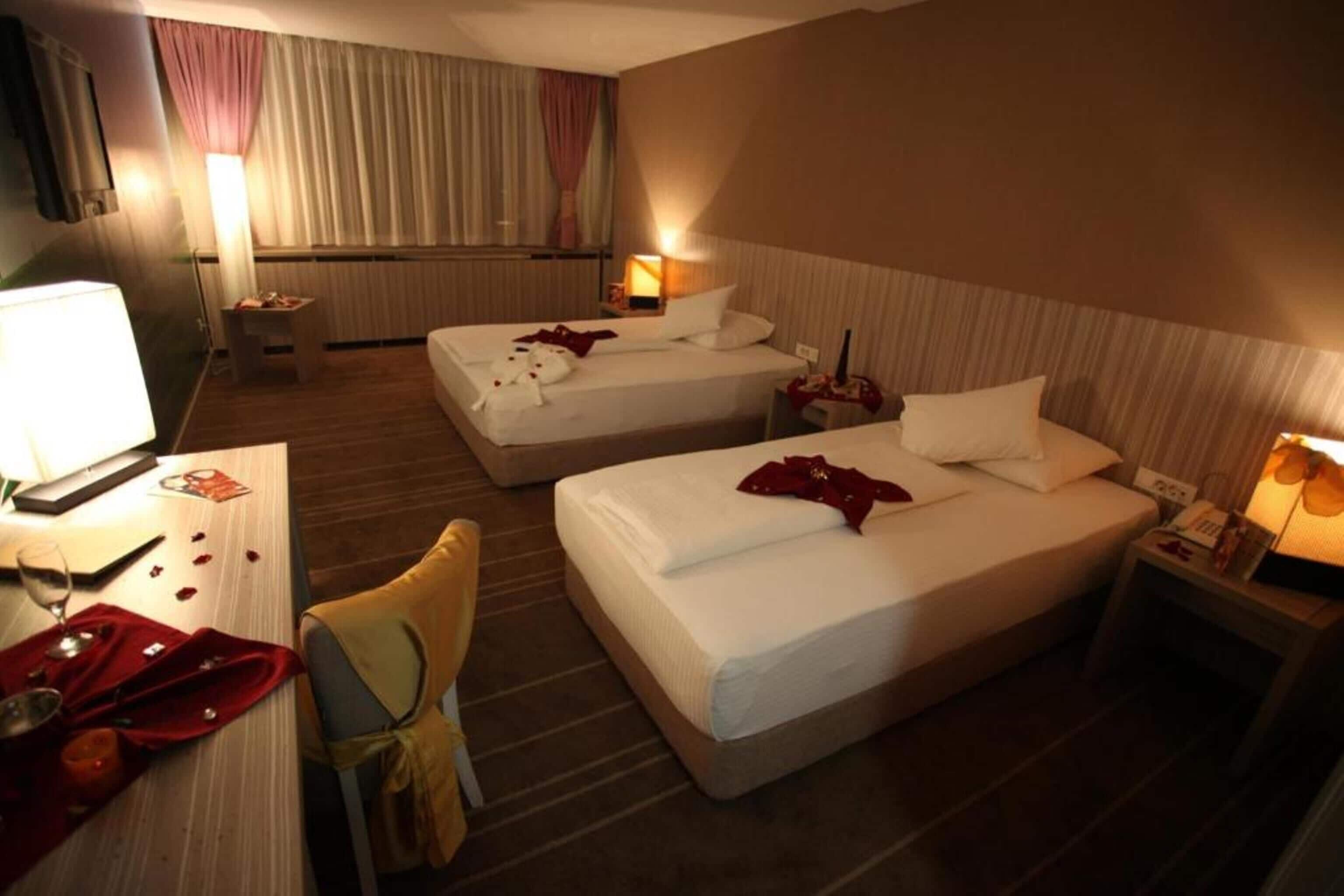 Quarto de hotel com duas camas, uma escrivaninha com computador, uma cadeira e mesa de cabeceira com abajur.