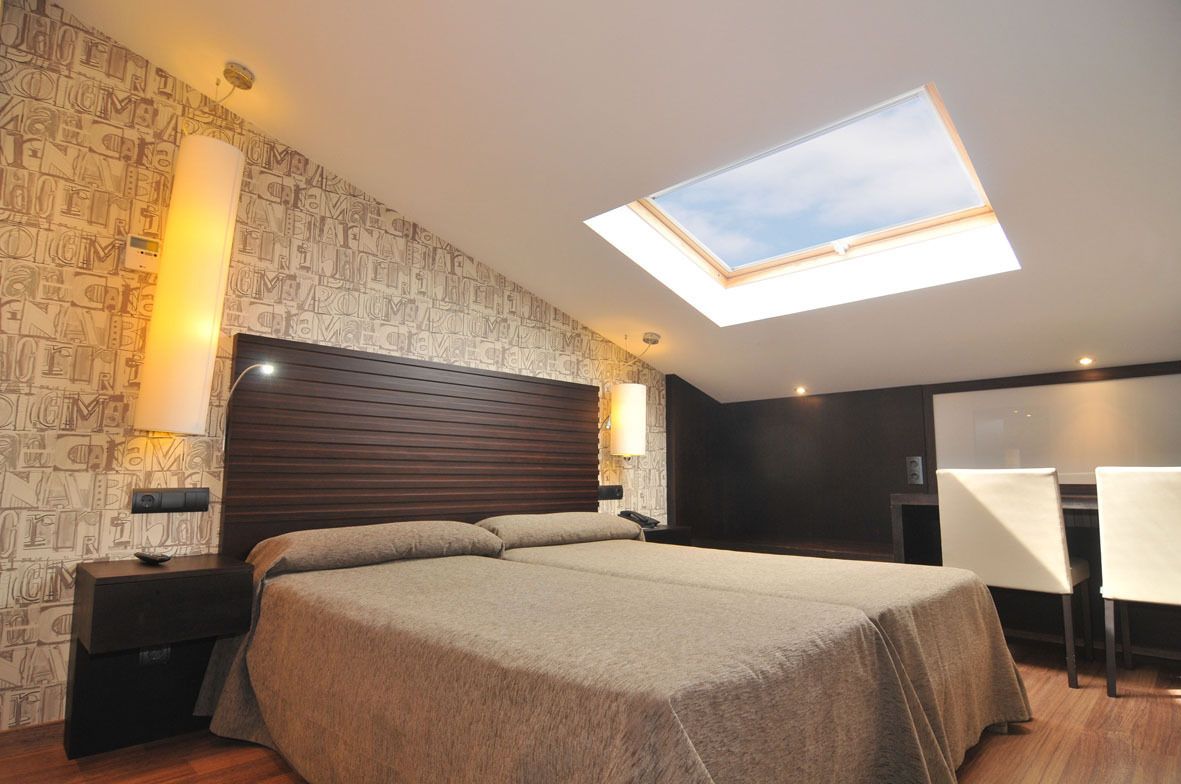 Quarto com claraboia, cama com cabeceira, luminárias de cabeceira e uma pequena mesa com cadeiras.
