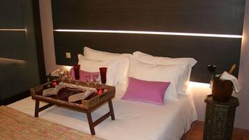 Quarto de hotel com uma cama bem arrumada, mesa de cabeceira com uma bandeja de copos de vinho e copos, e um vaso decorativo com uma rosa.