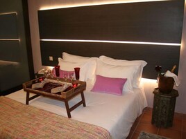 Quarto de hotel com uma cama bem arrumada, mesa de cabeceira com uma bandeja de copos de vinho e copos, e um vaso decorativo com uma rosa.