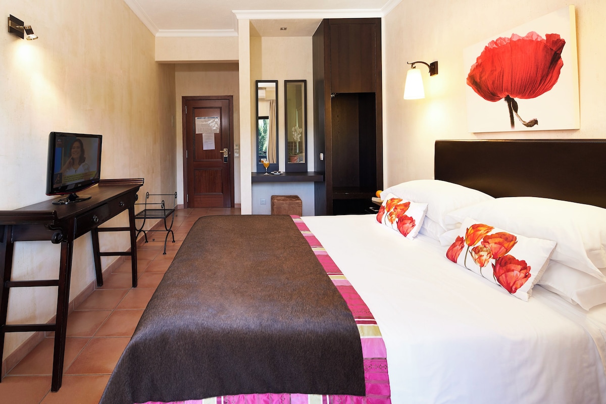 Quarto de hotel com uma cama, uma escrivaninha com computador, um espelho e um quadro de uma flor na parede.