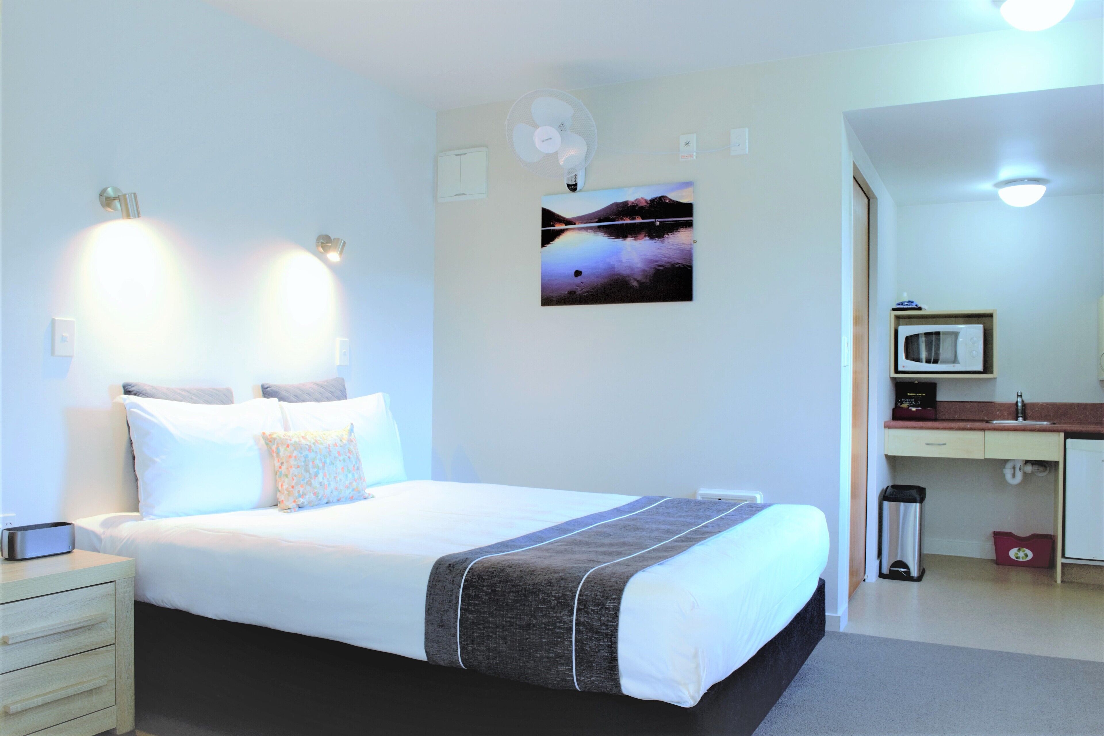 Quarto de hotel com cama, mesas de cabeceira, ventilador na parede, um quadro, micro-ondas e lixeira.