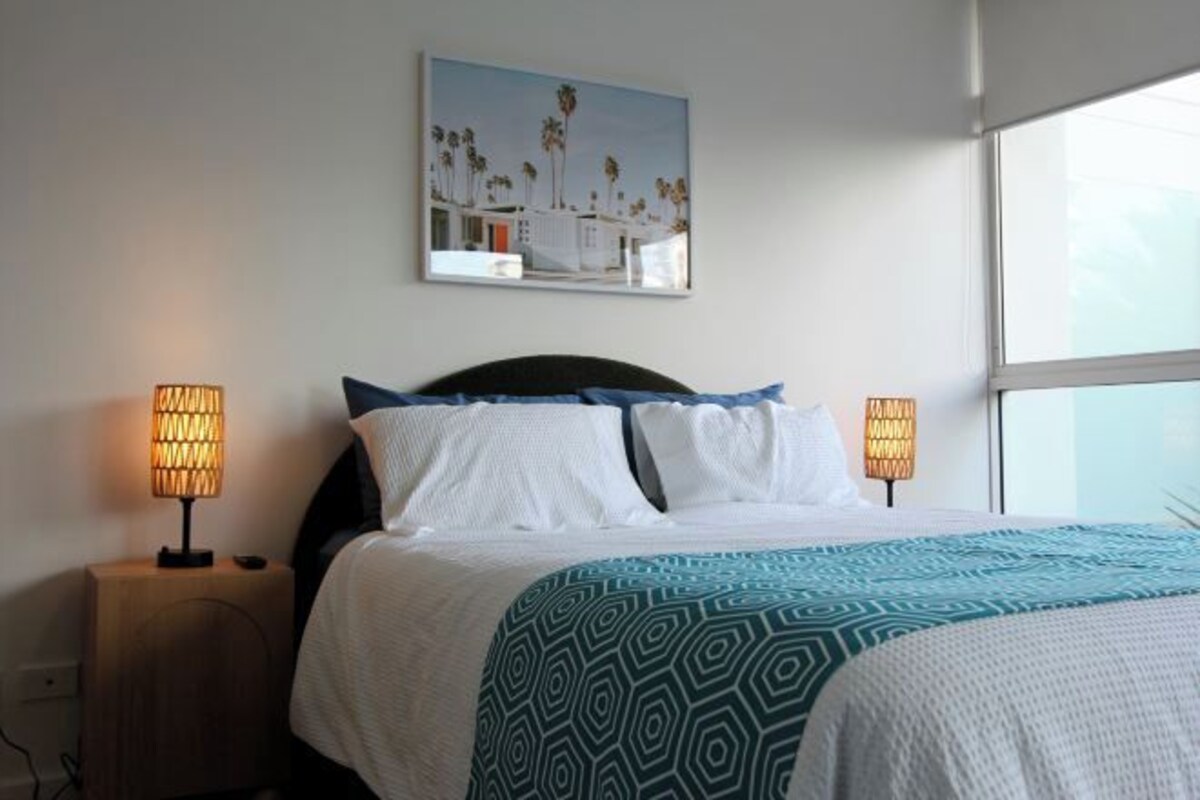 Quarto com cama, duas luminárias de cabeceira, mesa de cabeceira, janela e um quadro na parede.