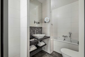 Uma casa de banho moderna com uma sanita branca, uma prateleira preta com toalhas, um espelho e uma banheira.