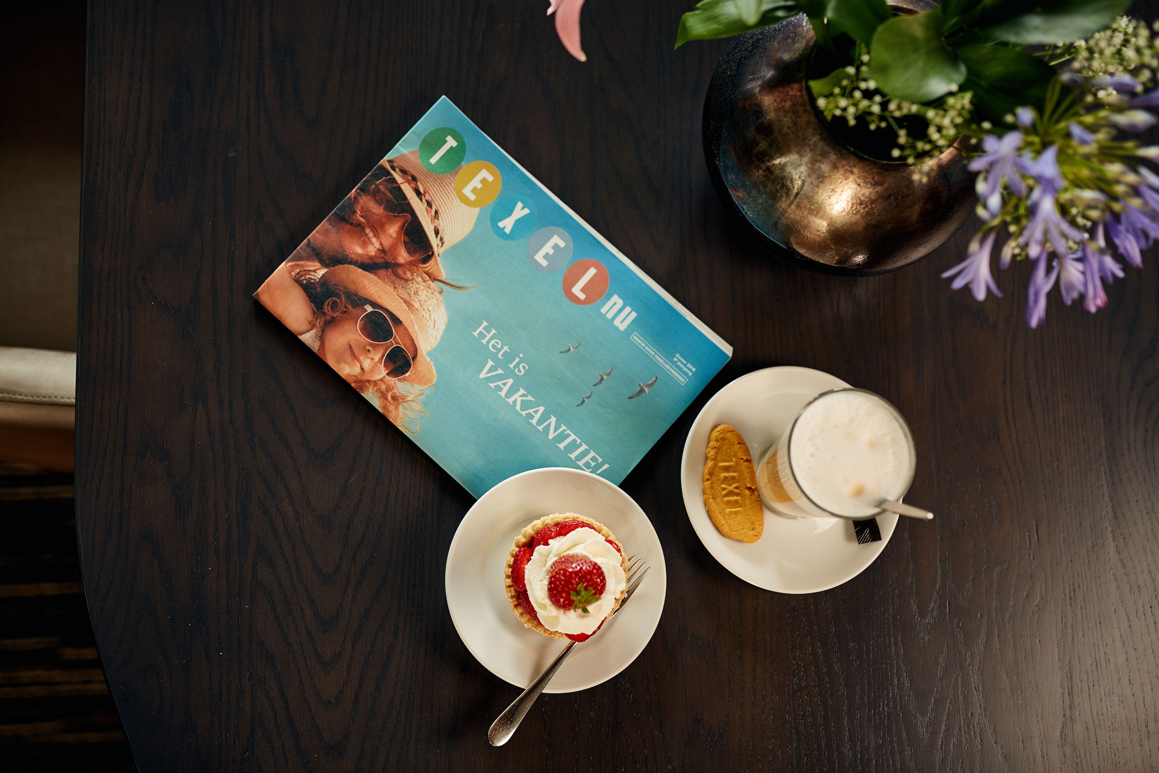 Um livro com o título "TEXTELN" sobre uma mesa de madeira, acompanhado de um prato com sobremesa e uma xícara de café.