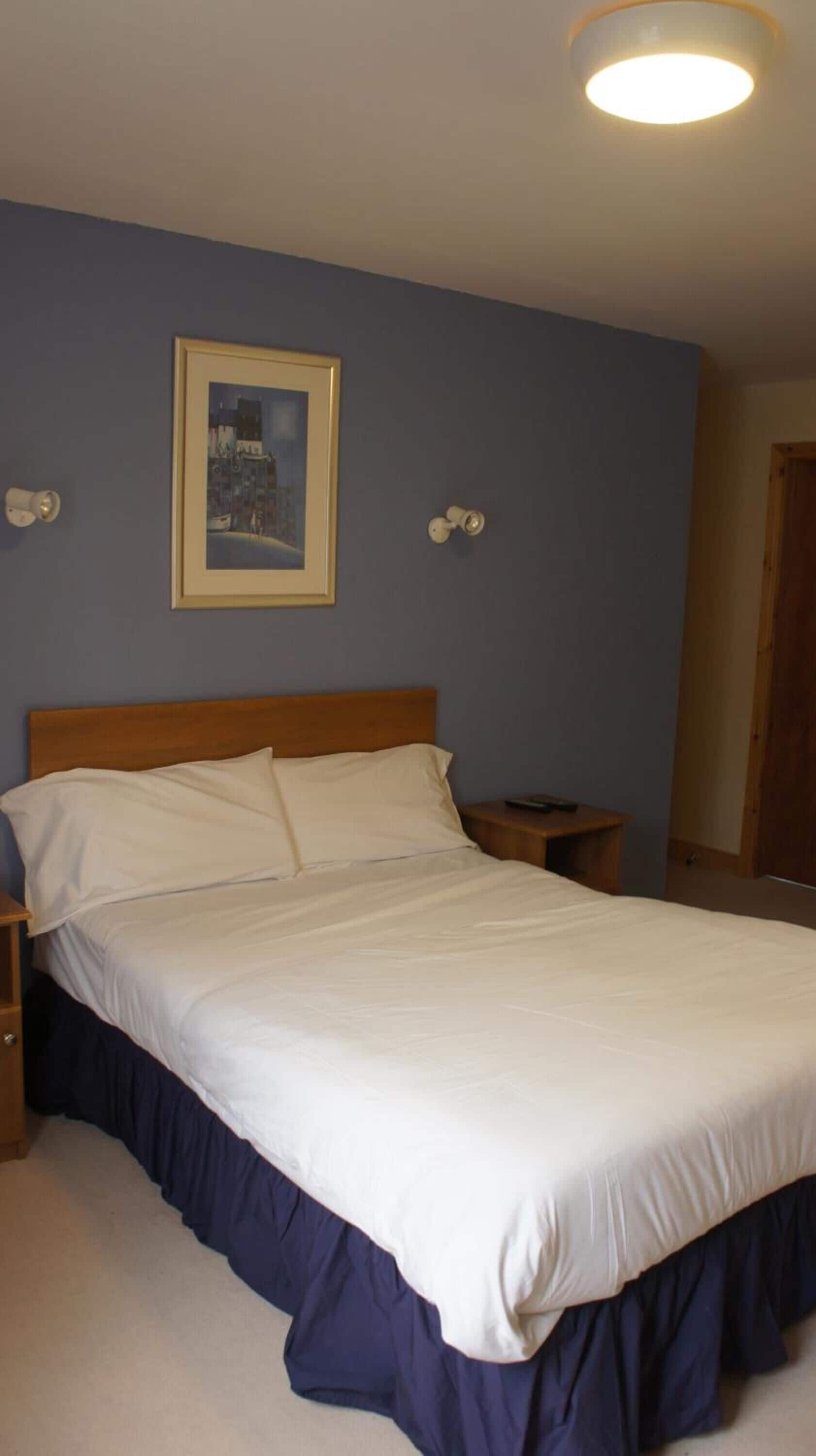 Cama bem arrumada com lençóis brancos, cabeceira de madeira e saia azul escura. Há um quadro emoldurado na parede acima da cama.