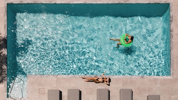 Uma piscina retangular com água cristalina, uma pessoa flutuando em um círculo inflável verde e outra deitada à beira da piscina.