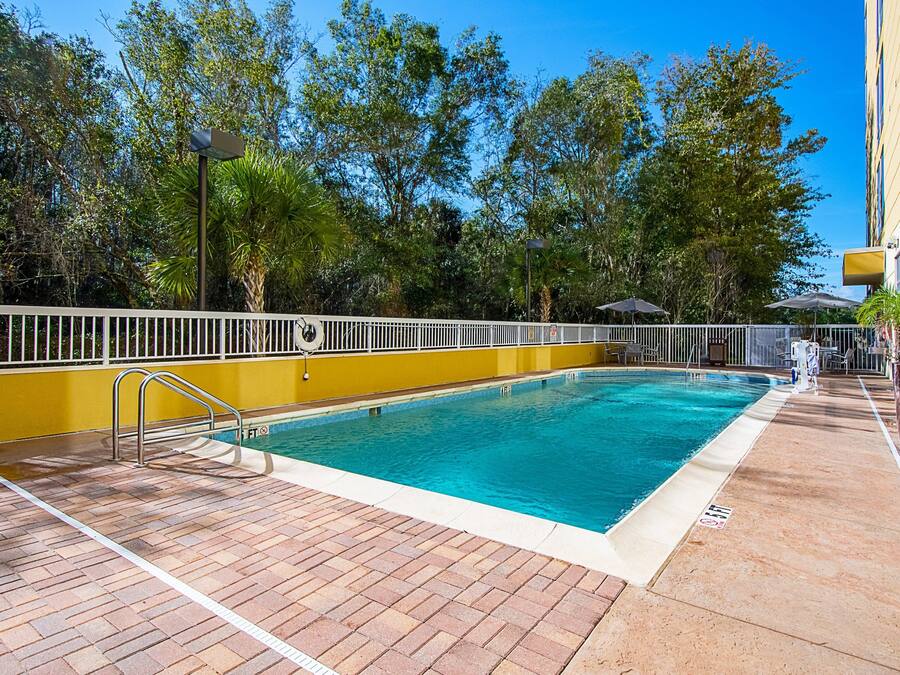 Piscina retangular com um deck branco, cercada por uma grade branca, e árvores ao fundo.