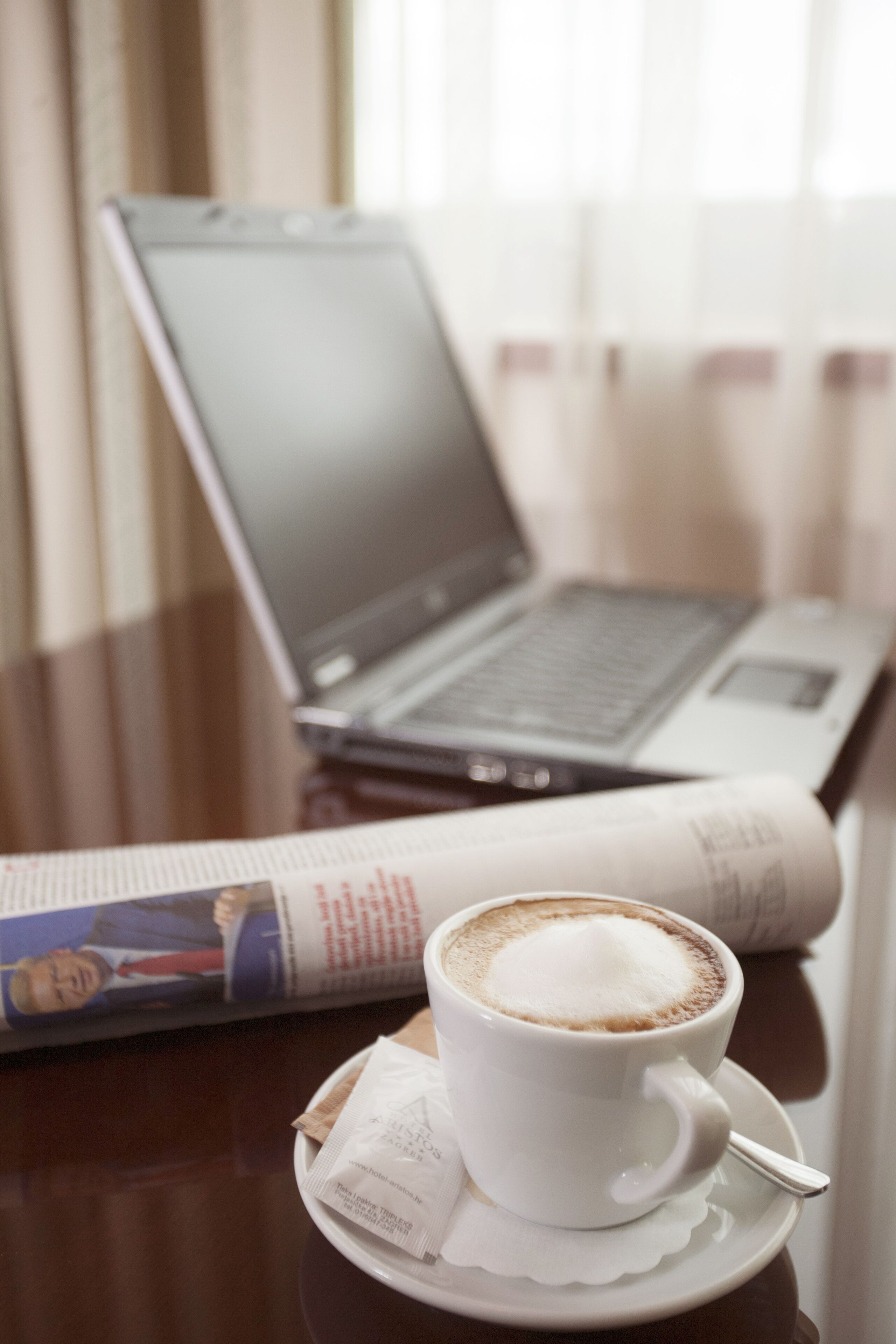 Um laptop sobre uma mesa com uma chávena de café e um jornal.