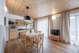 Cozinha moderna com armários de madeira, uma mesa de jantar e cadeiras. Há uma televisão em um suporte e uma janela com cortinas.
