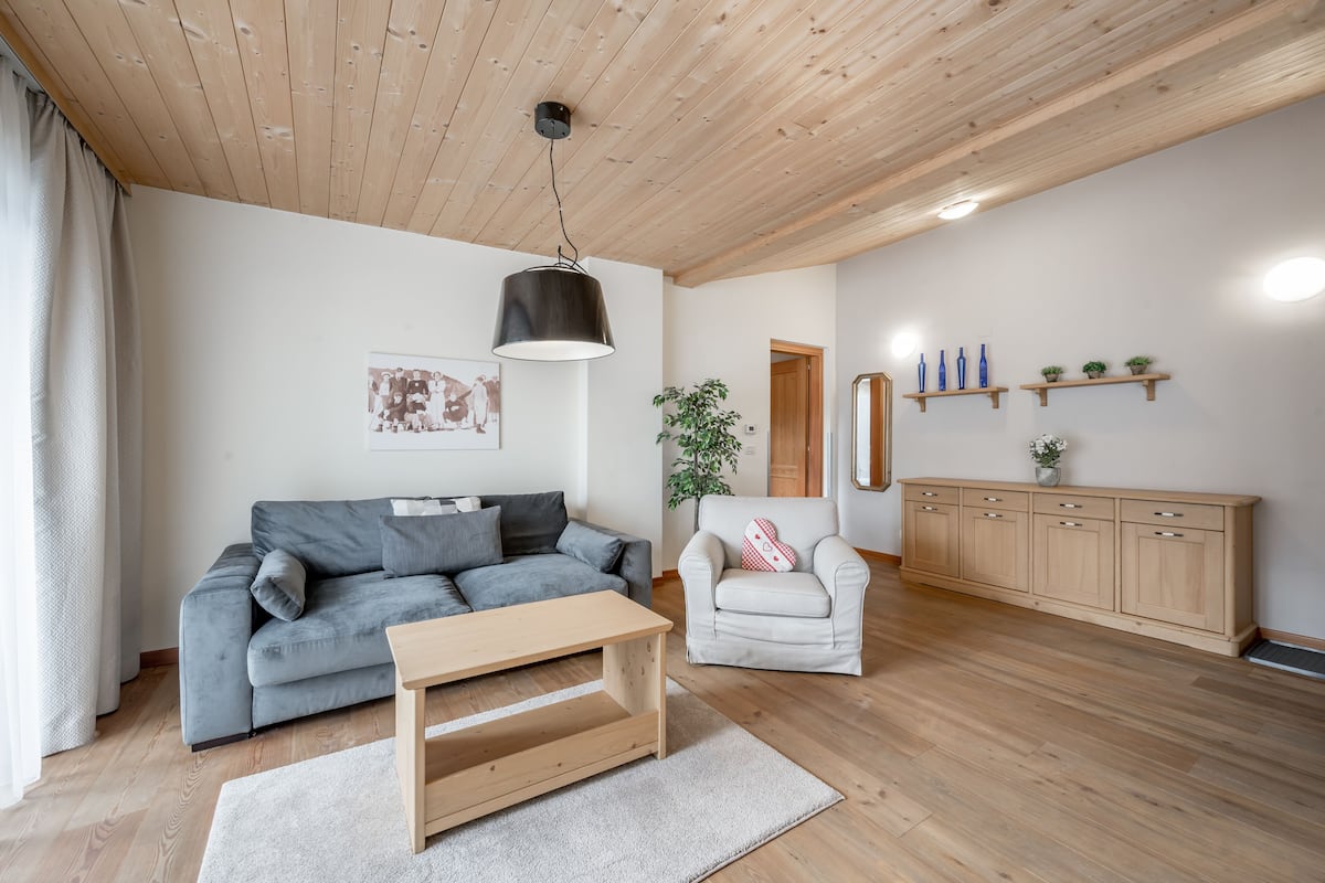 Uma sala de estar moderna com um sofá cinzento, uma mesa de centro de madeira, uma poltrona branca e um armário de madeira com prateleiras.