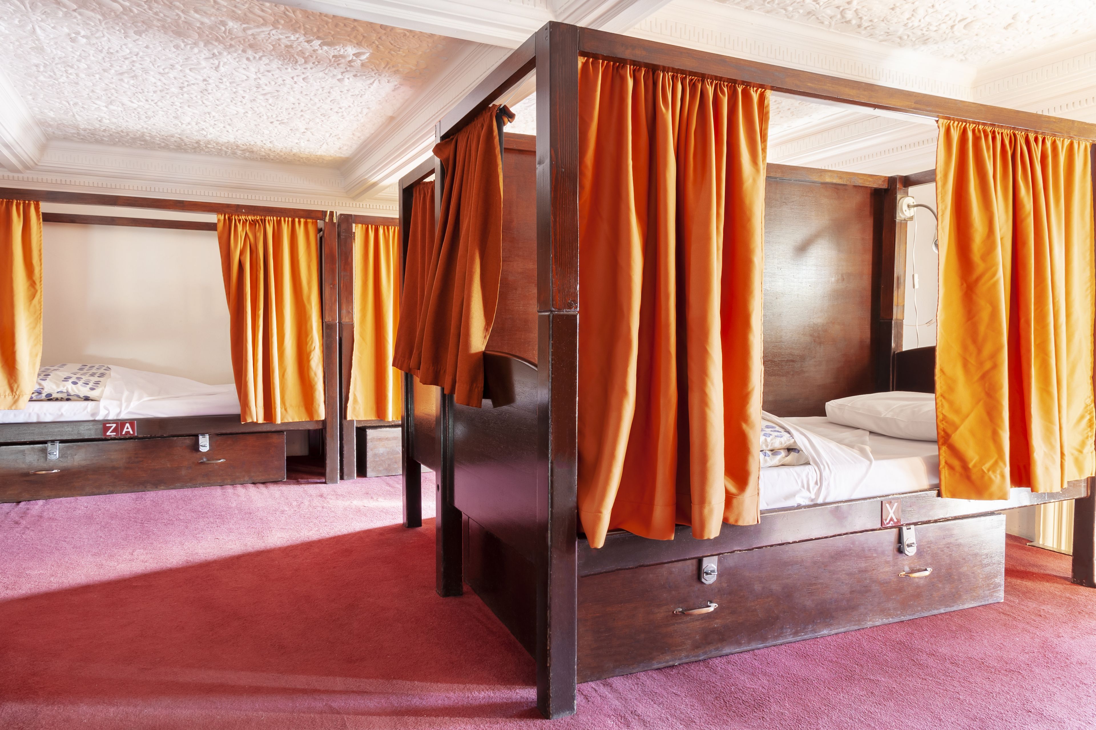 Quarto de hotel com quatro camas de solteiro, cada uma com estrutura de madeira e cortinas laranja.
