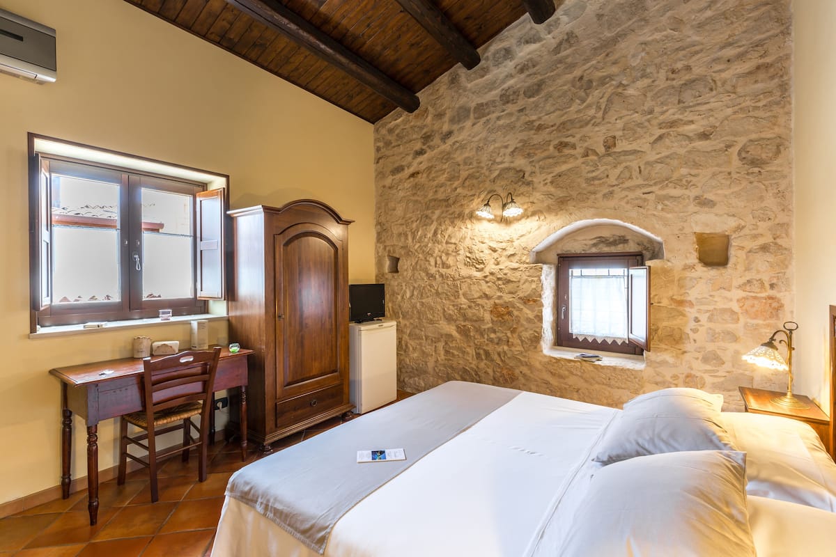 Quarto com parede de pedra, escrivaninha de madeira, cadeira, cama e mesa de cabeceira com abajur.