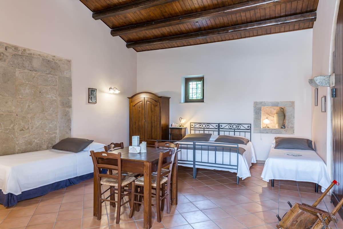 Quarto com duas camas de solteiro, uma mesa de jantar de madeira com cadeiras, uma parede de pedra, um guarda-roupa de madeira e uma pequena janela.