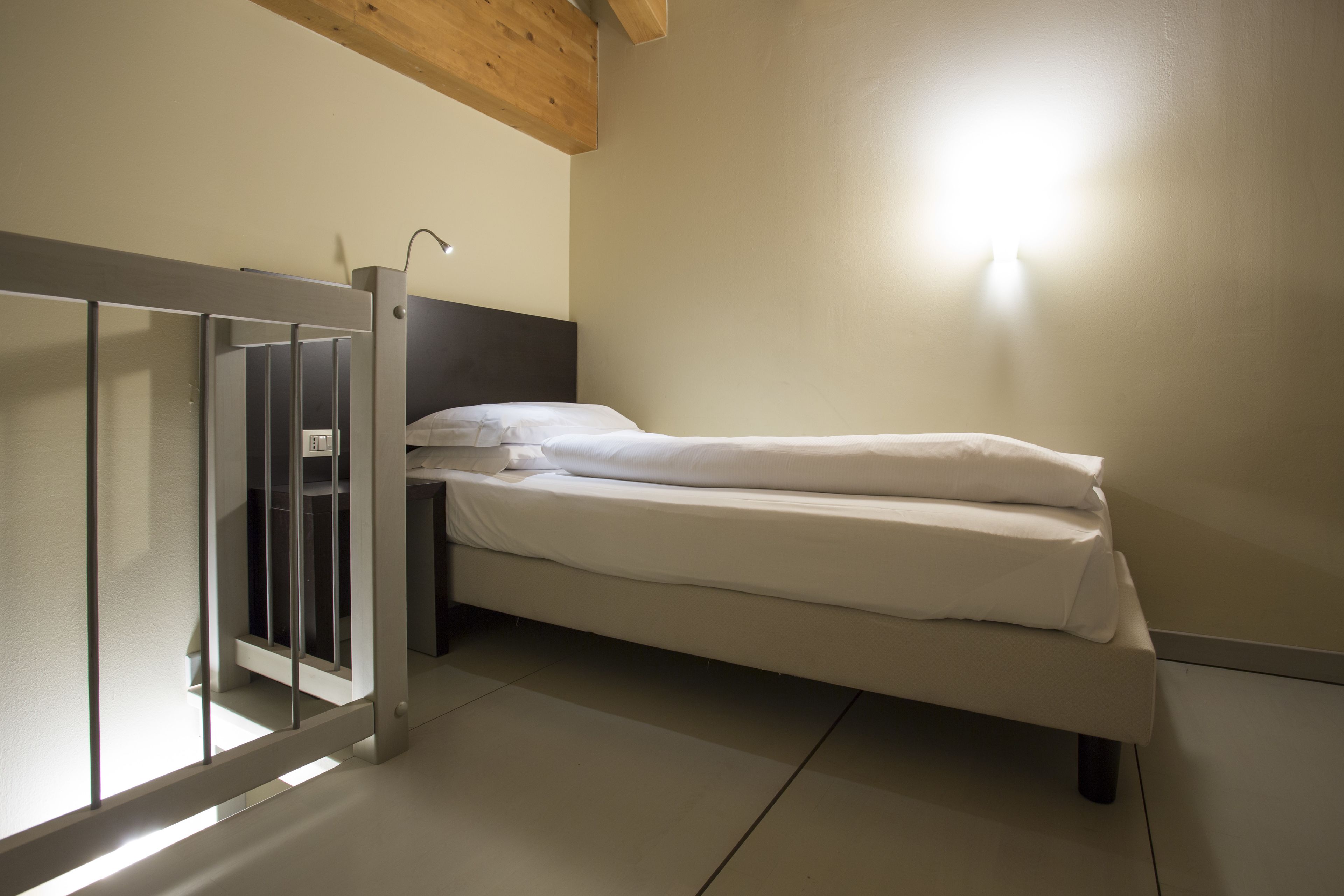 Uma cama de solteiro num quarto com vigamento de madeira no teto, cabeceira branca e uma fonte de luz na parede.