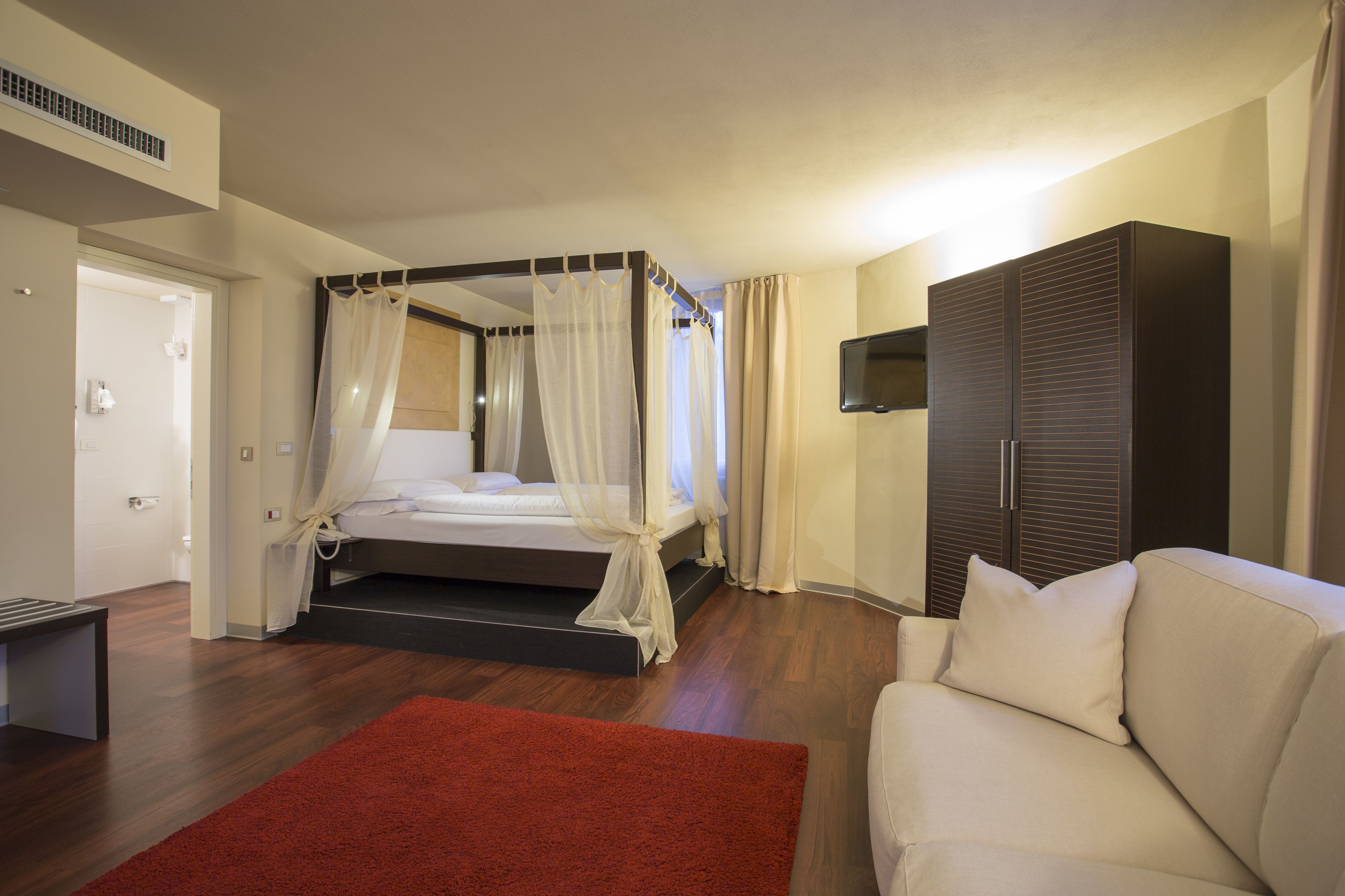 Quarto com cama de dossel, sofá, televisão fixada na parede e guarda-roupa.
