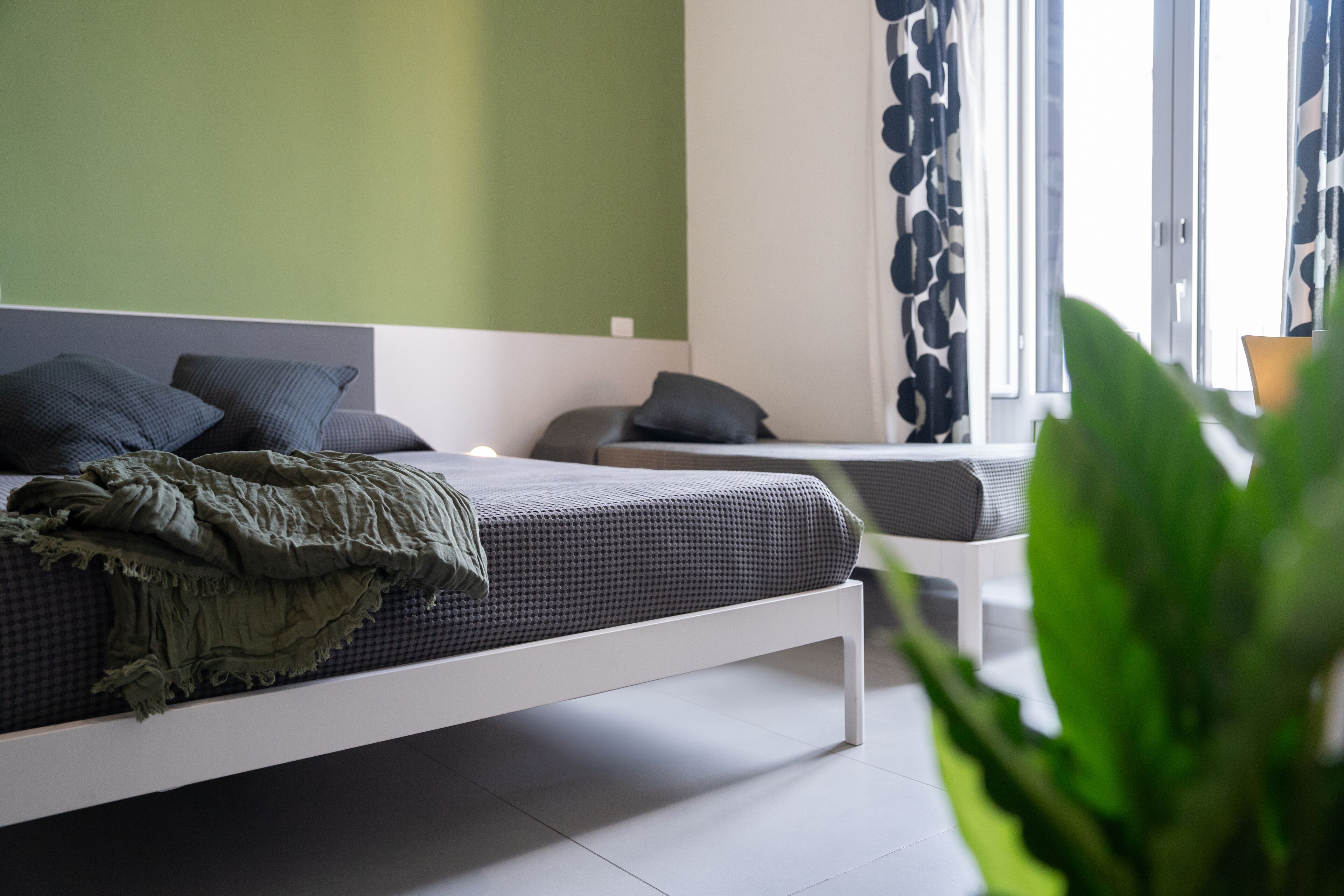 Quarto moderno com duas camas de solteiro, parede verde e uma janela grande com cortinas.