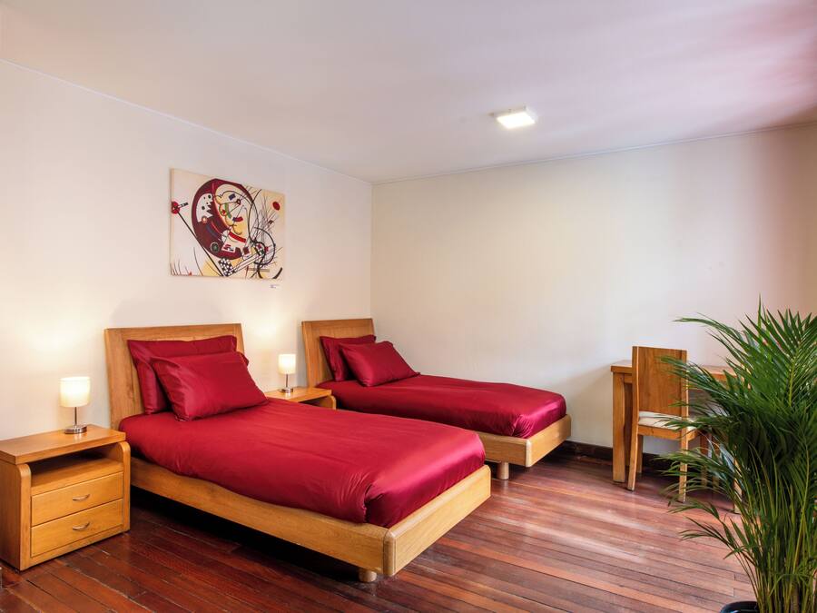 Quarto com duas camas de madeira, cada uma com roupa de cama vermelha, um criado-mudo com abajur, uma cadeira de madeira e uma grande planta em vaso.