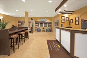 Um saguão de hotel com balcão de recepção, área de bar com banquetas, espaço de estar com cadeiras e uma grande janela com cortinas.