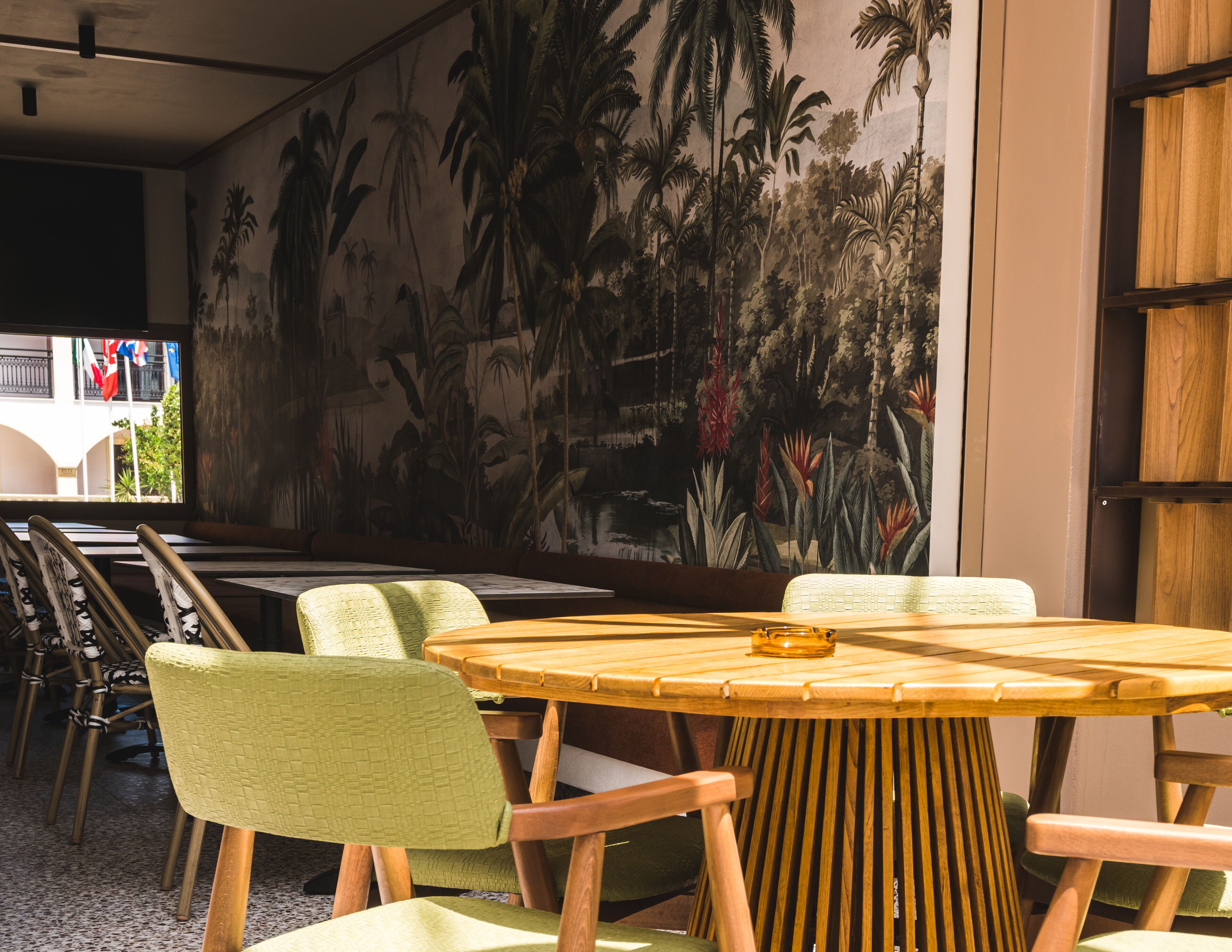 Interior de restaurante com um mural de plantas tropicais, mesas e cadeiras de madeira, e vista para o exterior através de uma janela.
