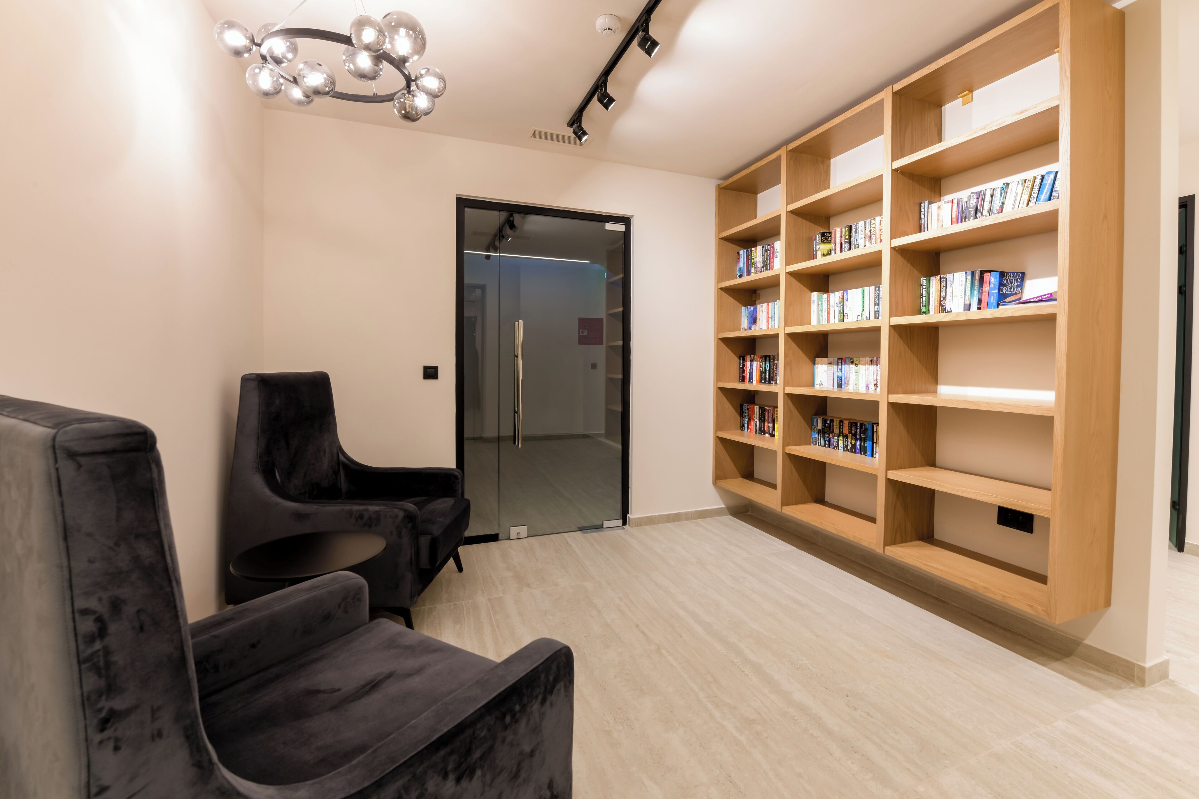 Uma sala de leitura moderna com duas cadeiras pretas, uma estante de madeira cheia de livros e uma porta de vidro que dá para outro espaço.