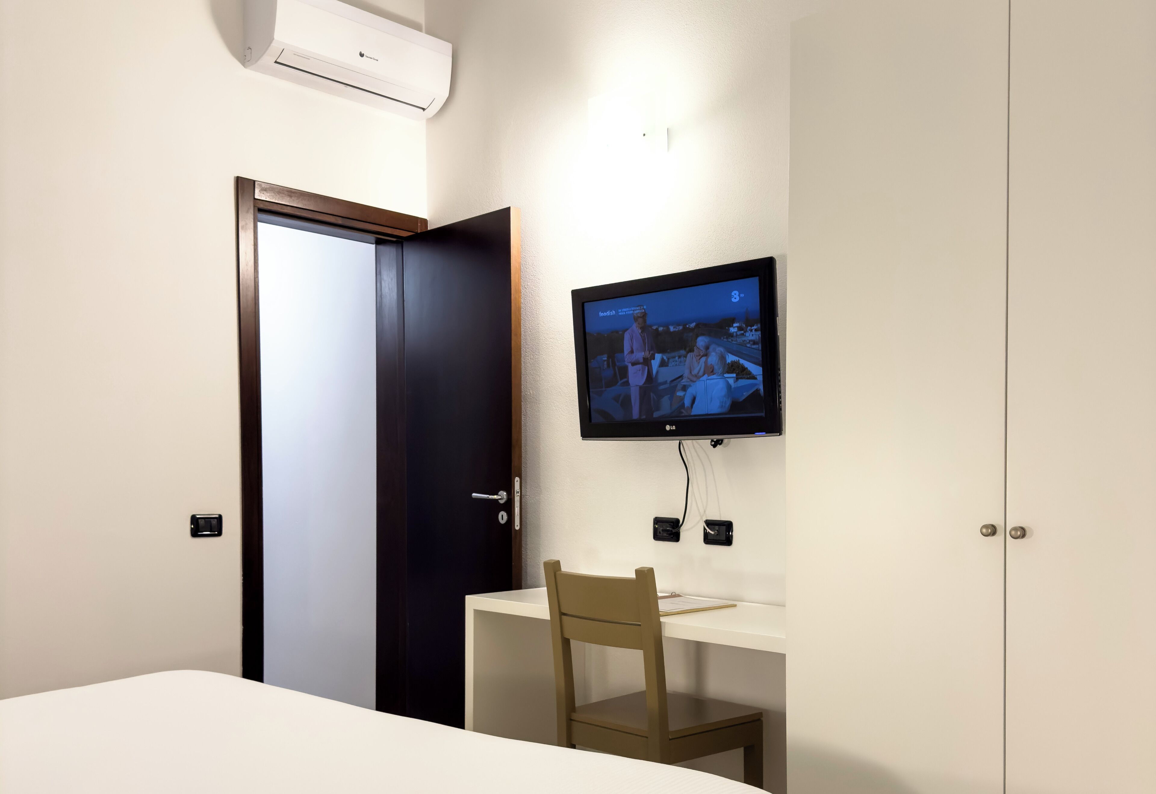 Quarto de hotel com cama, escrivaninha com cadeira, televisão na parede e porta.