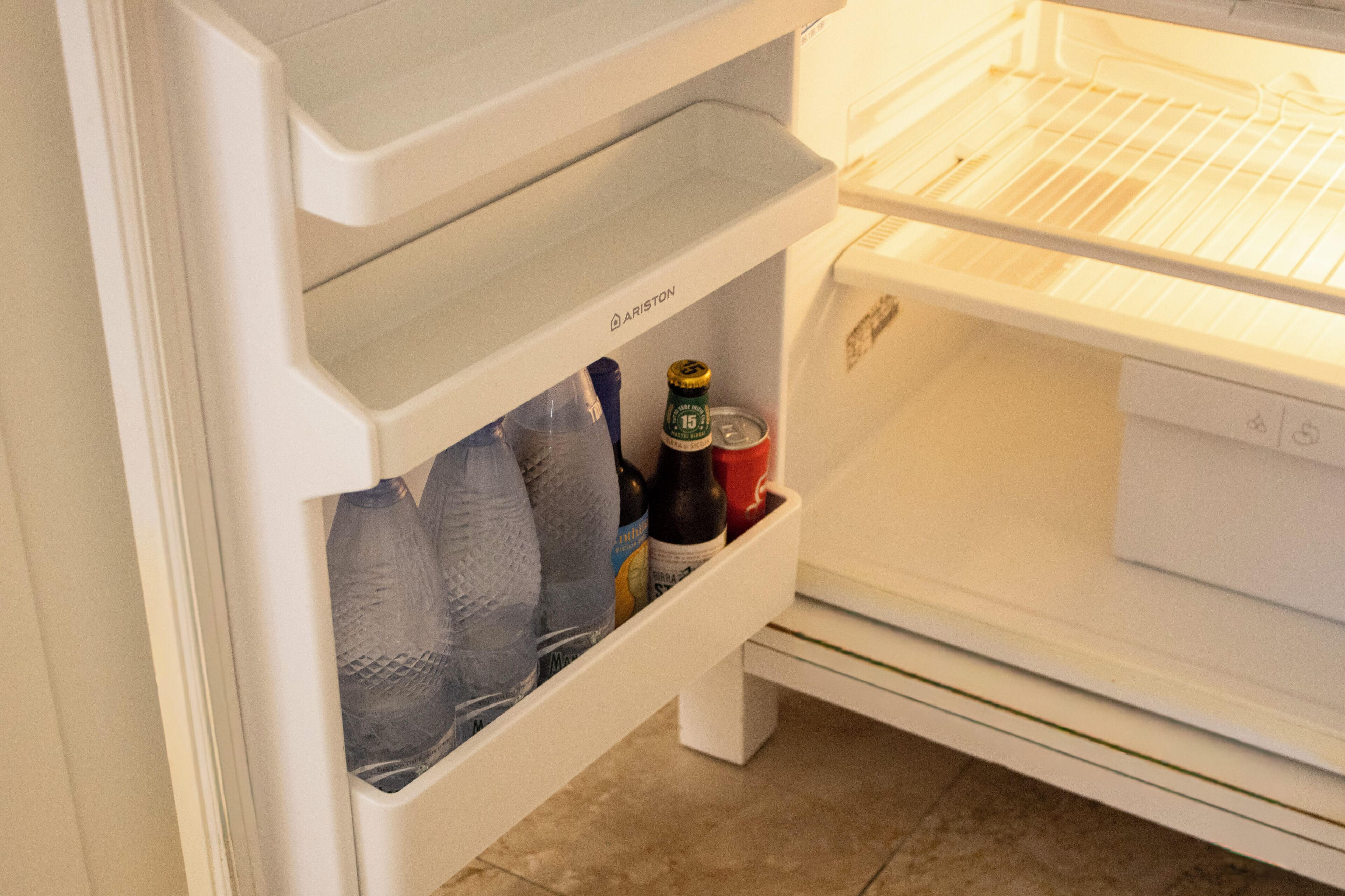 Uma geladeira branca com compartimento para congelador, contendo uma garrafa de cerveja, uma lata de refrigerante e um saco de compras.
